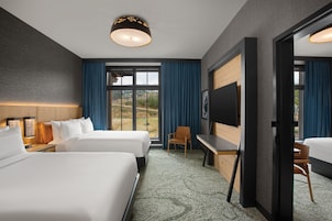 Quarto de hotel com duas camas, uma escrivaninha, uma cadeira, uma televisão e uma janela grande com cortinas.