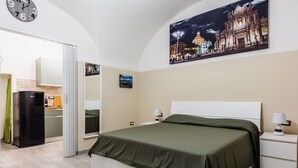 Quarto com cama, mesas de cabeceira, relógio na parede, pintura e vista para uma cozinha.