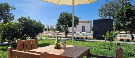 Uma mesa de madeira com um grande guarda-sol, rodeada por vegetação exuberante e uma área de piscina ao fundo.