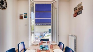 Uma sala de jantar com mesa de madeira posta para quatro, cadeiras e vista para uma varanda com toldo azul.