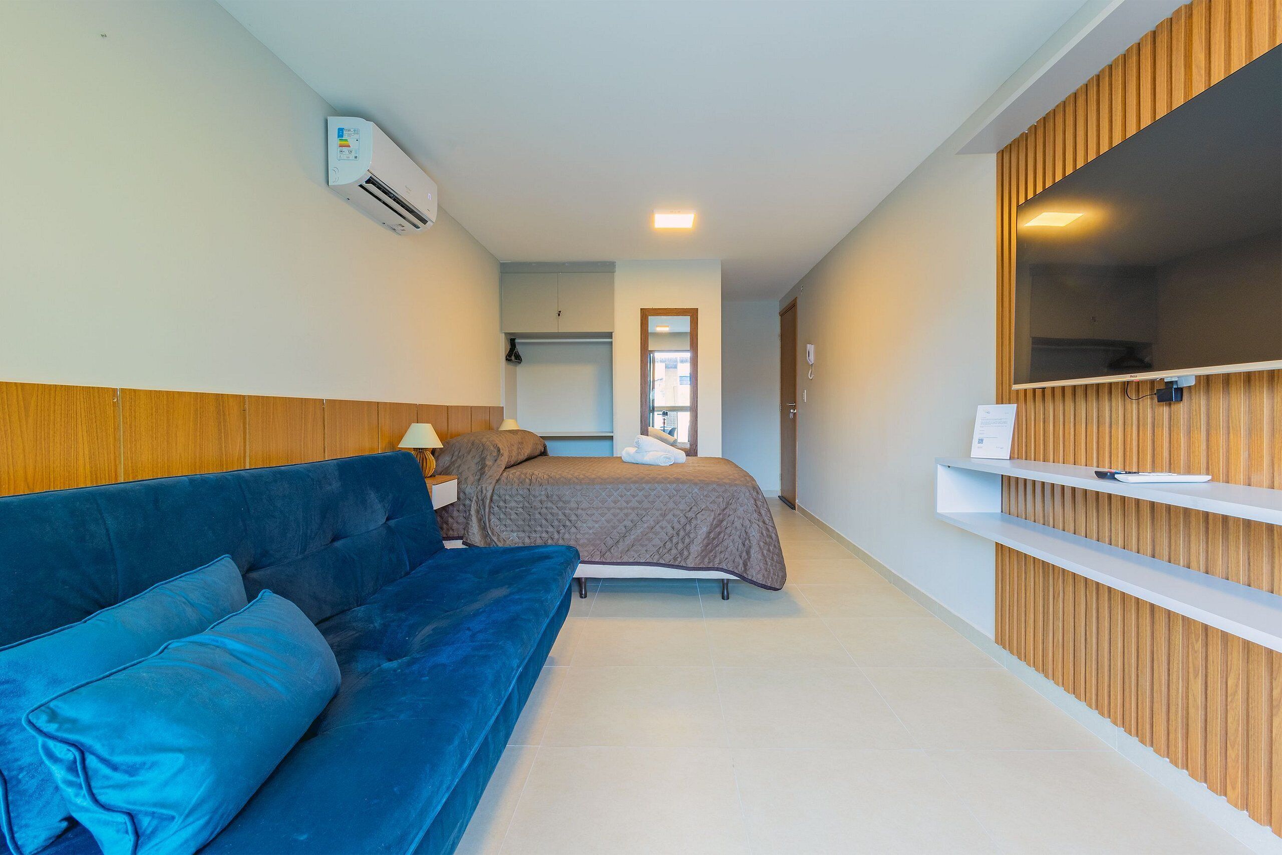 Quarto com sofá azul, cama com manta marrom, parede de painéis de madeira e prateleira branca.