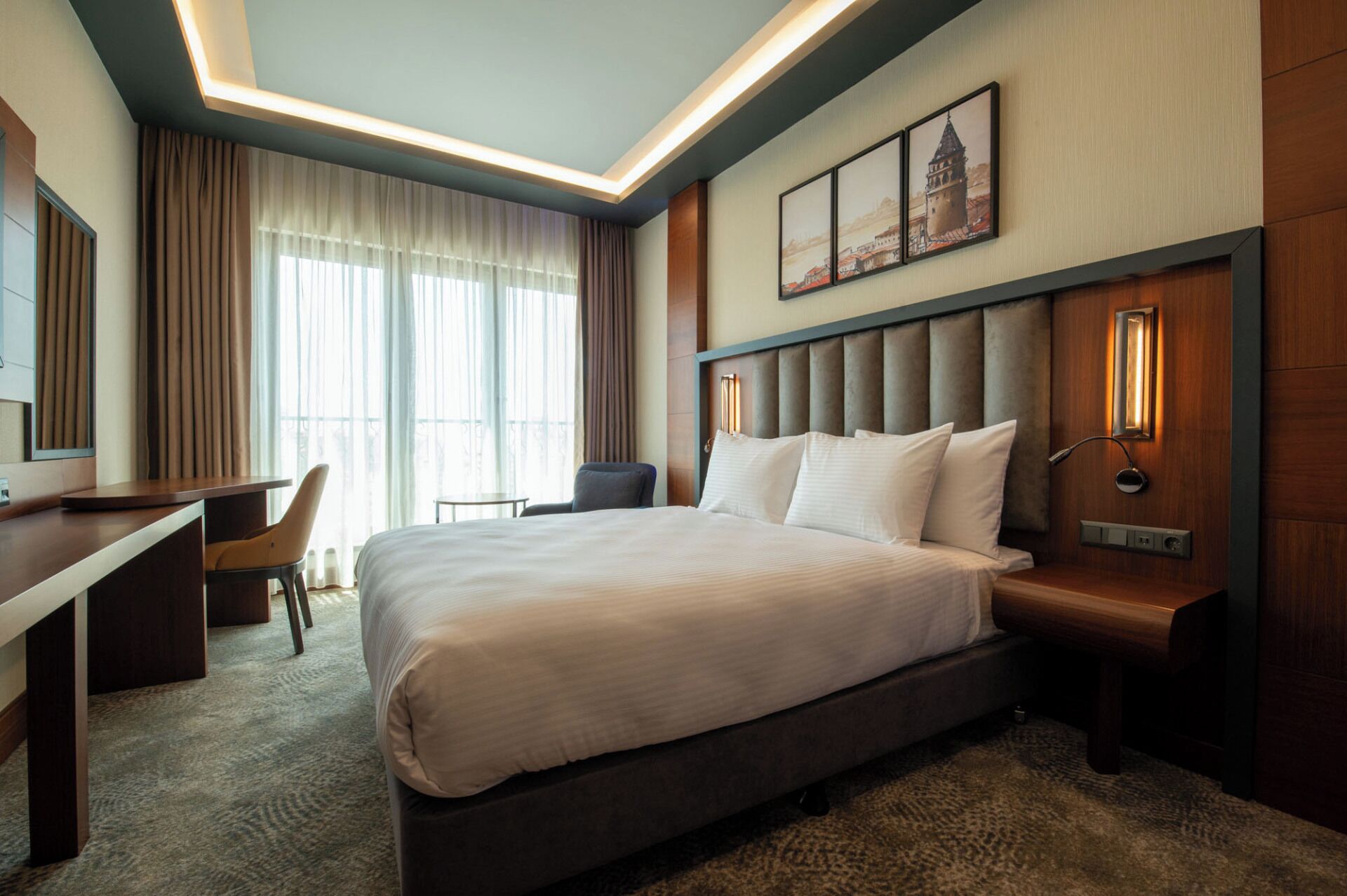 Quarto de hotel com uma cama grande, uma escrivaninha, uma cadeira e vista para a cidade pela janela.