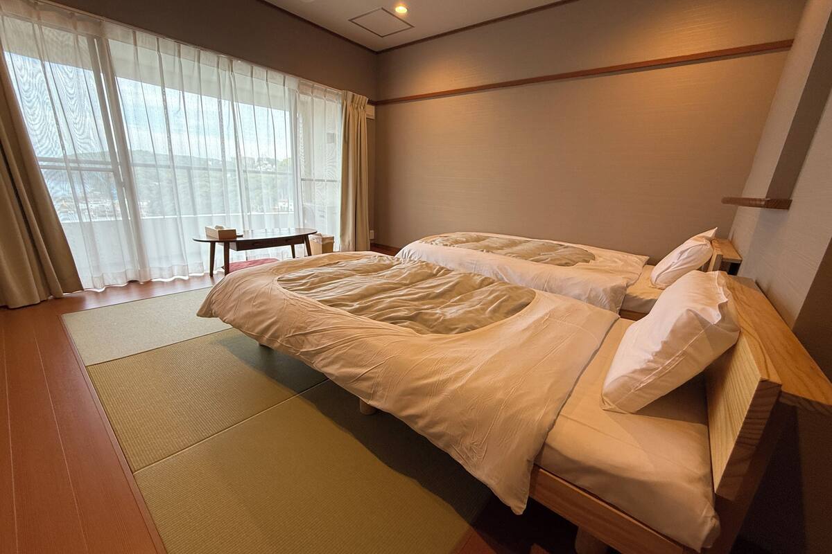 Quarto de hotel com duas camas, uma mesa pequena e vista através de cortinas finas.