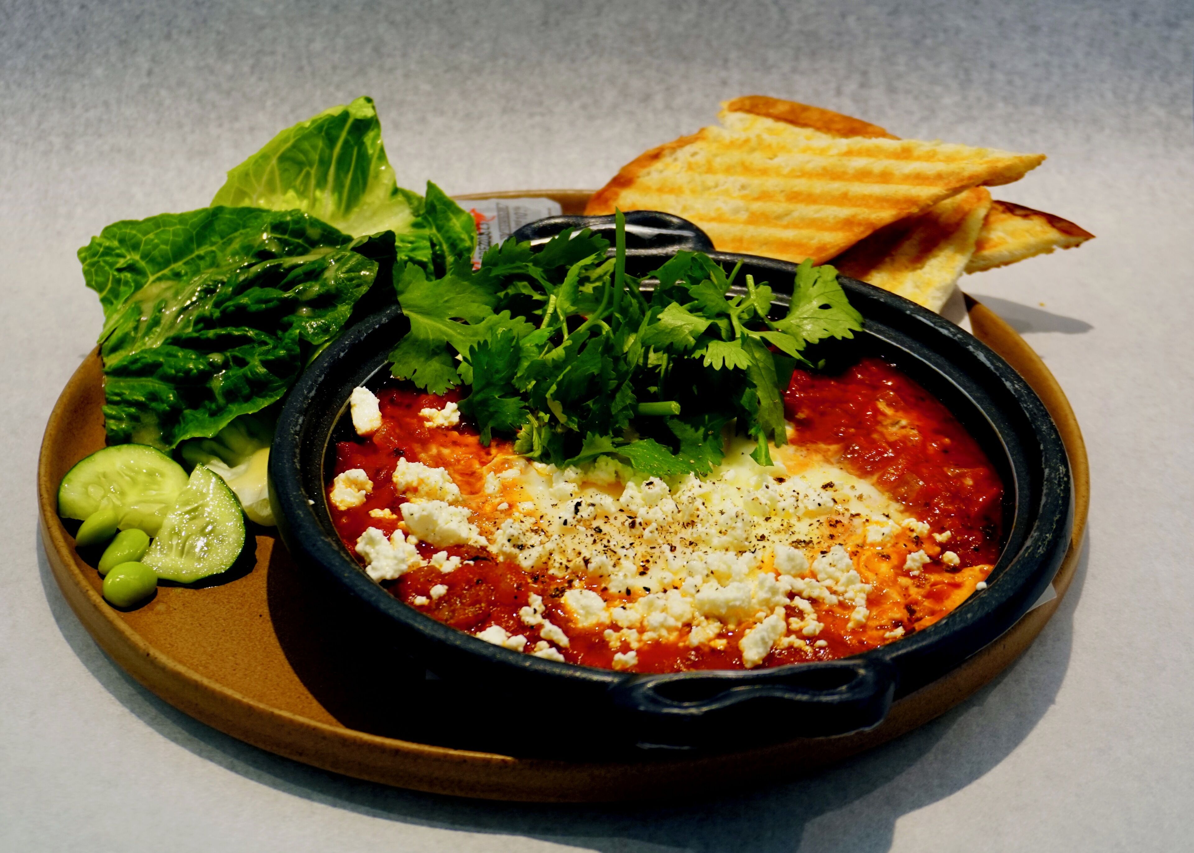 Uma tigela com prato à base de tomate, queijo feta, guarnecida com ervas, acompanhada de pão grelhado e legumes frescos.