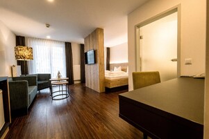 Quarto de hotel com piso de madeira, um sofá, uma mesinha, uma cama e uma televisão fixada na parede.