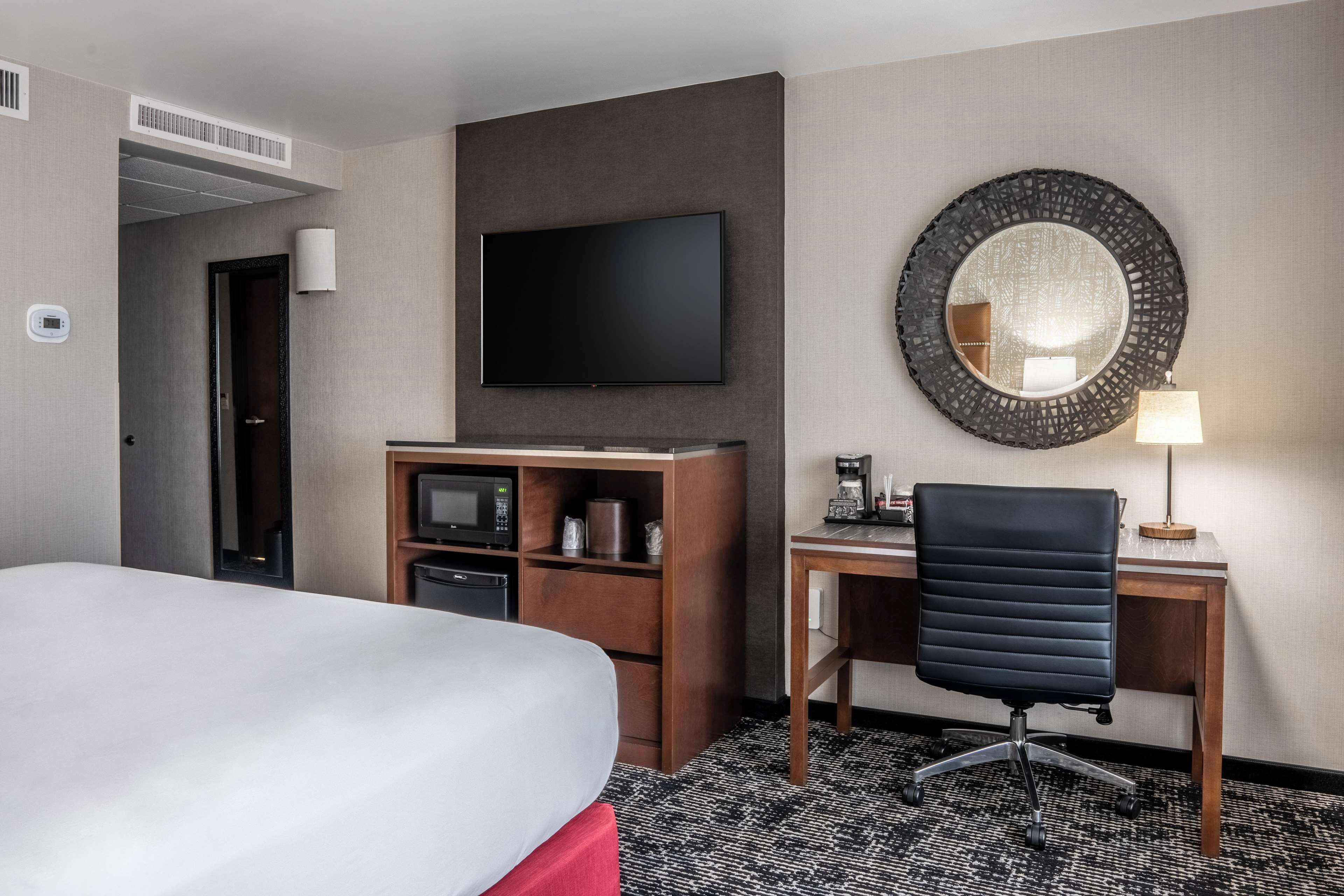 Quarto de hotel com cama, escrivaninha e cadeira, televisão, máquina de café, micro-ondas e um espelho grande.