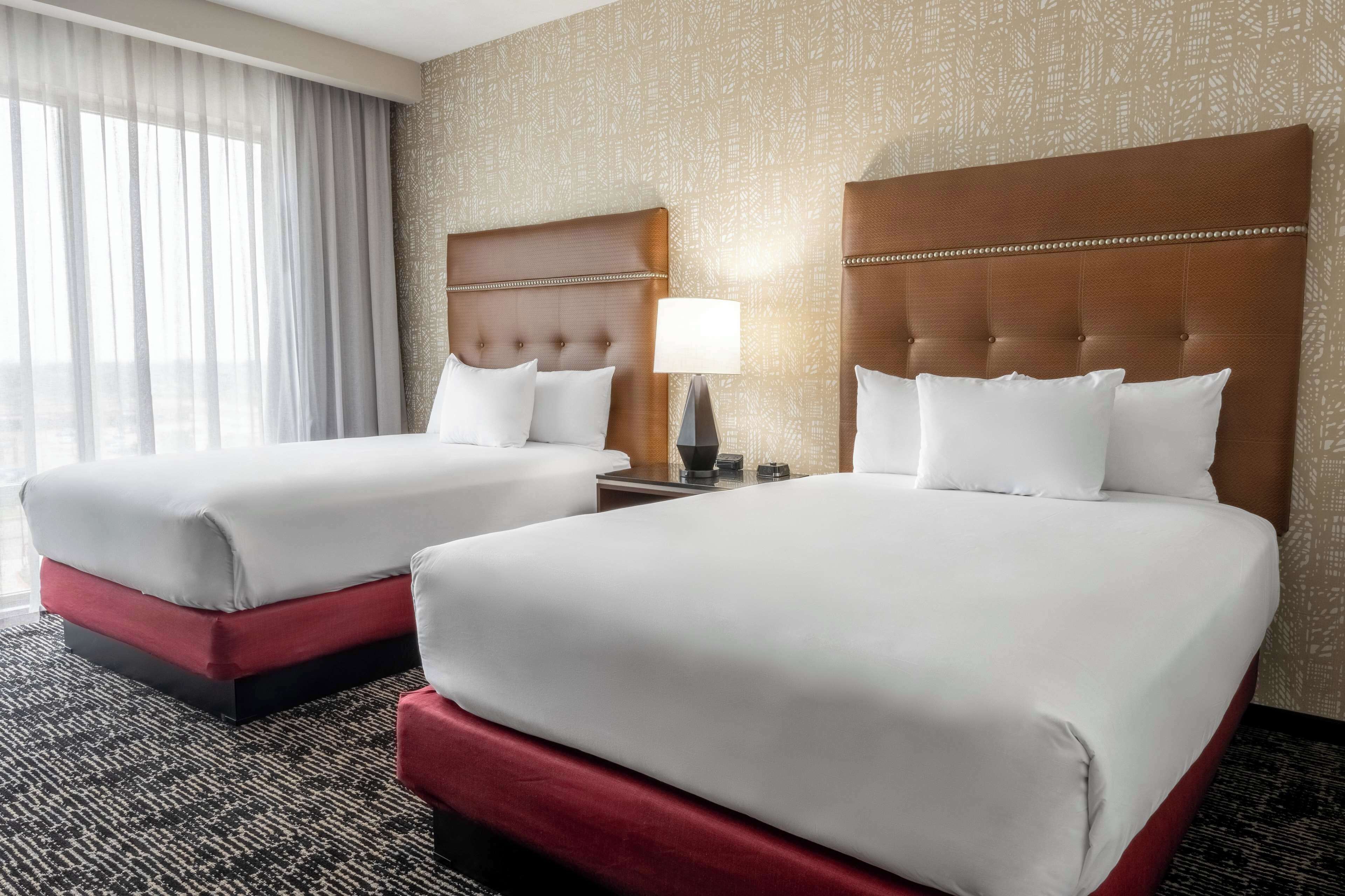 Quarto de hotel com duas camas, uma janela grande com cortinas translúcidas, parede estampada e mesa de cabeceira com abajur.