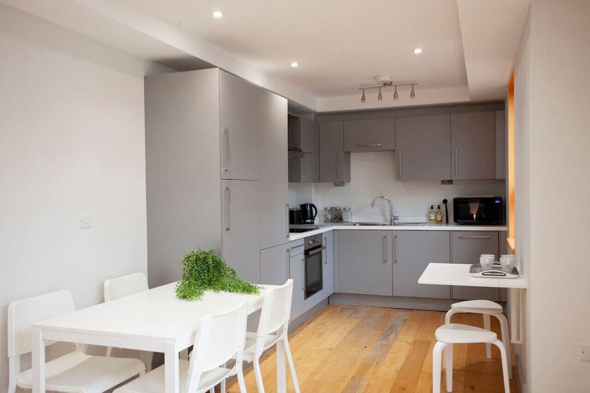 Cozinha moderna com armários cinza, bancada branca e um pequeno espaço de refeições com mesa e cadeiras brancas.