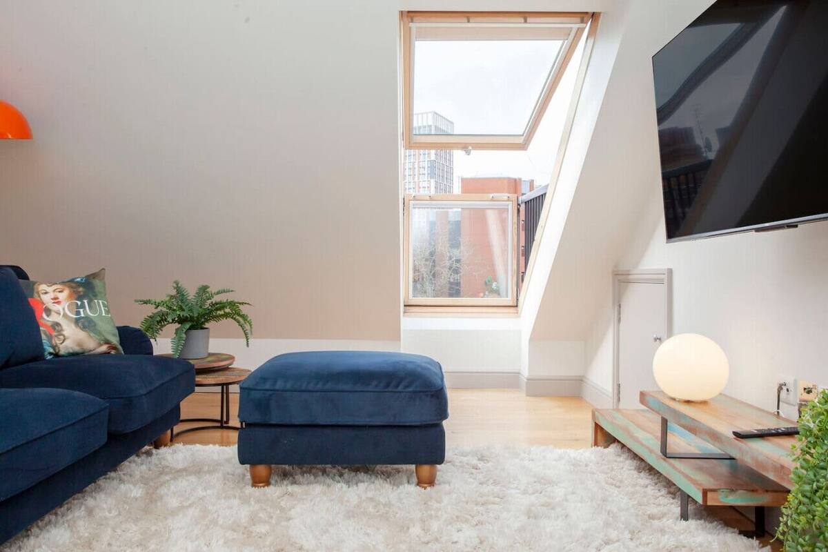Uma sala de estar moderna com um sofá azul, um tapete branco e fofo, uma mesinha redonda com uma planta, e uma janela grande com vista para a cidade.