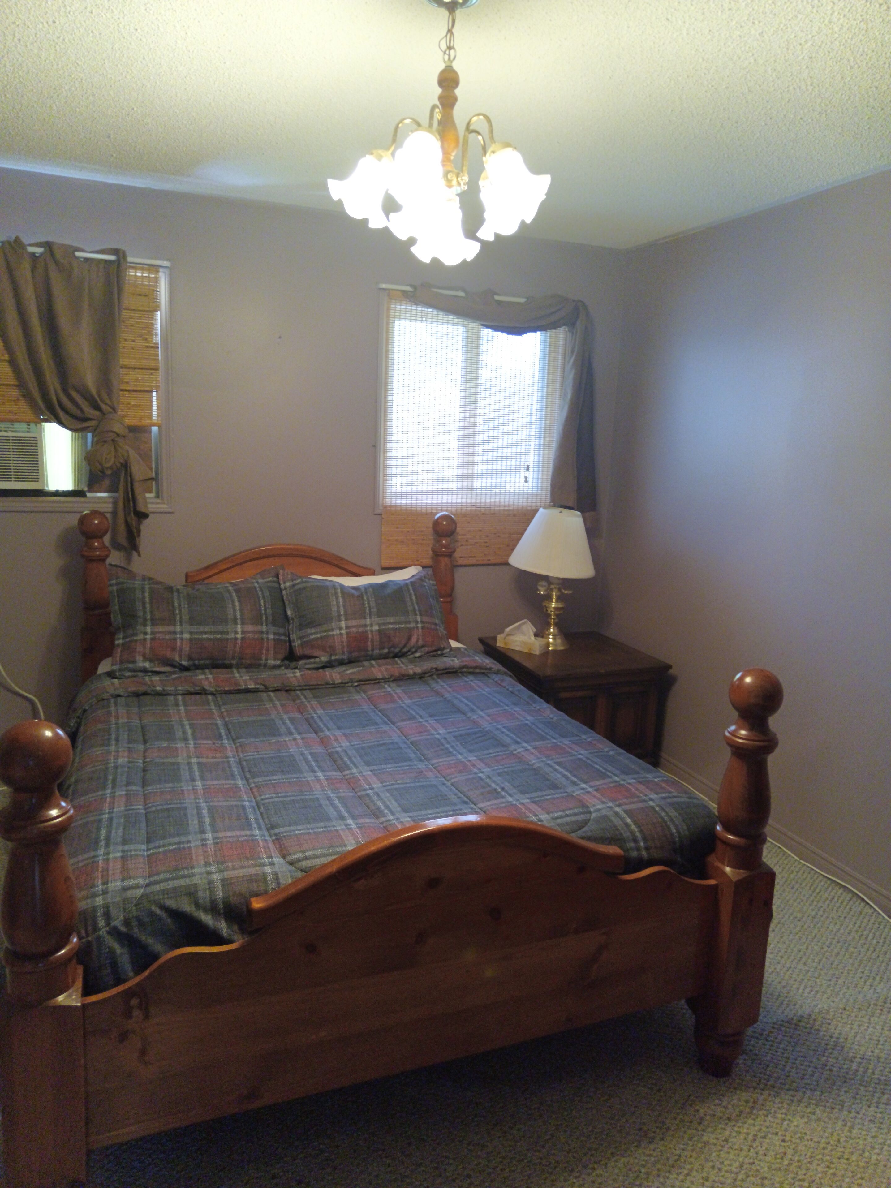 Cama bem arrumada com colcha xadrez e almofadas, cabeceira de madeira, mesa de cabeceira com abajur e janela com persianas.