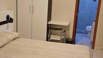Quarto com cama, escrivaninha e cadeira. Um banheiro está visível por uma porta aberta.