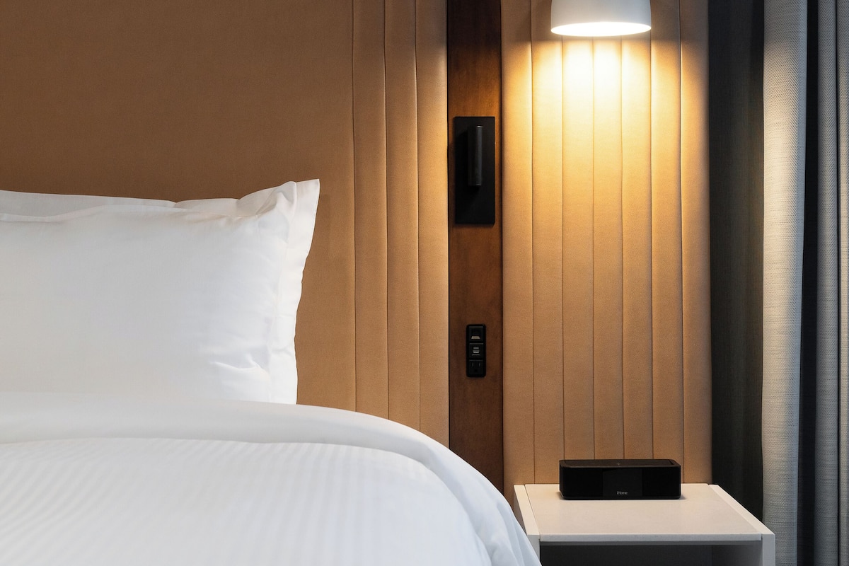 Quarto de hotel com uma cama grande, uma mesa de cabeceira com abajur e um painel de controle fixado na parede.