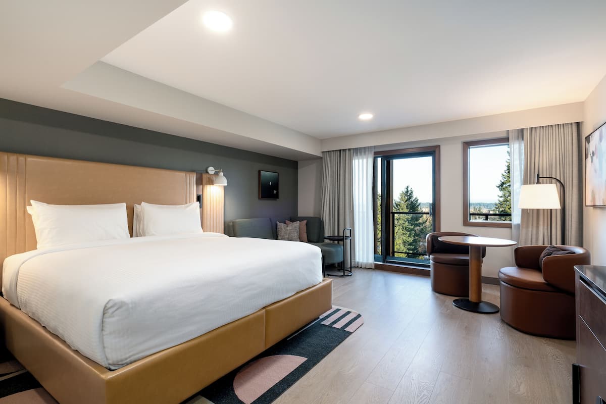 Quarto de hotel moderno com uma cama grande, uma escrivaninha, duas cadeiras, uma mesinha e vista para o exterior.