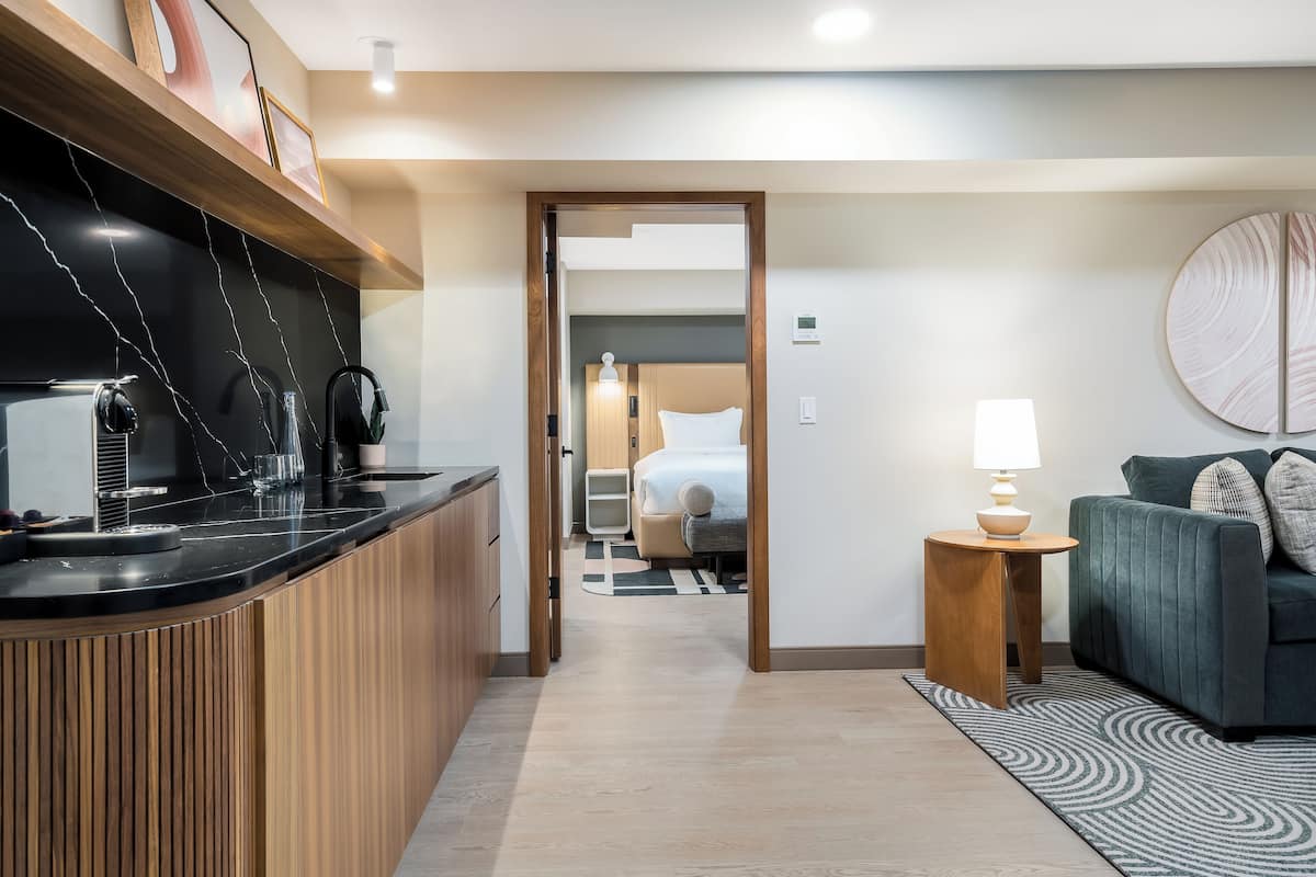 Quarto de hotel moderno com cozinha, sofá, luminária e cama com mesa de cabeceira.