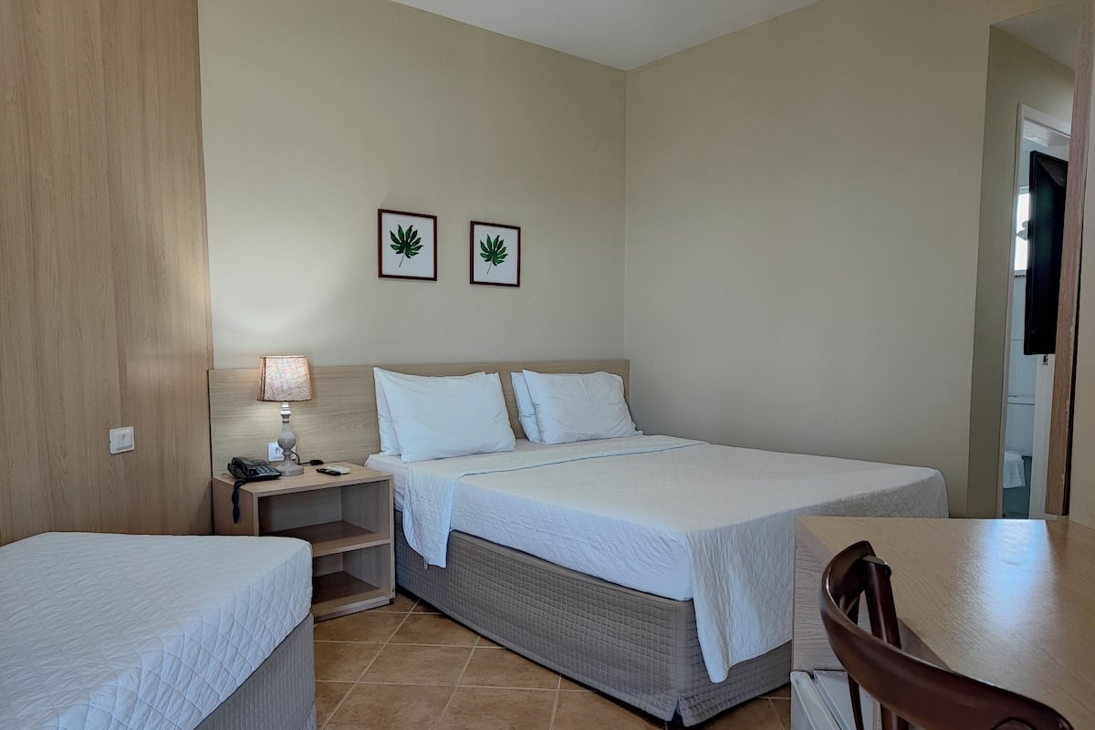 Quarto de hotel com duas camas, uma escrivaninha, uma cadeira, uma mesa de cabeceira com abajur e duas quadros na parede.