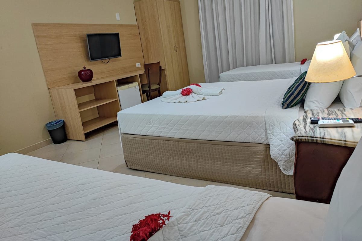 Quarto de hotel com duas camas, uma TV fixada num móvel de madeira, uma escrivaninha com luminária e um cesto de lixo.