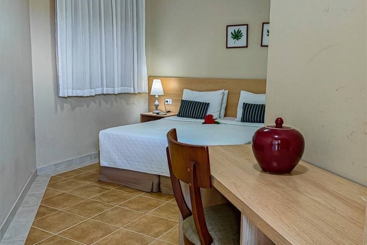 Quarto de hotel com uma cama, uma mesa de cabeceira, uma luminária, uma cadeira e um vaso vermelho sobre uma mesa de madeira.