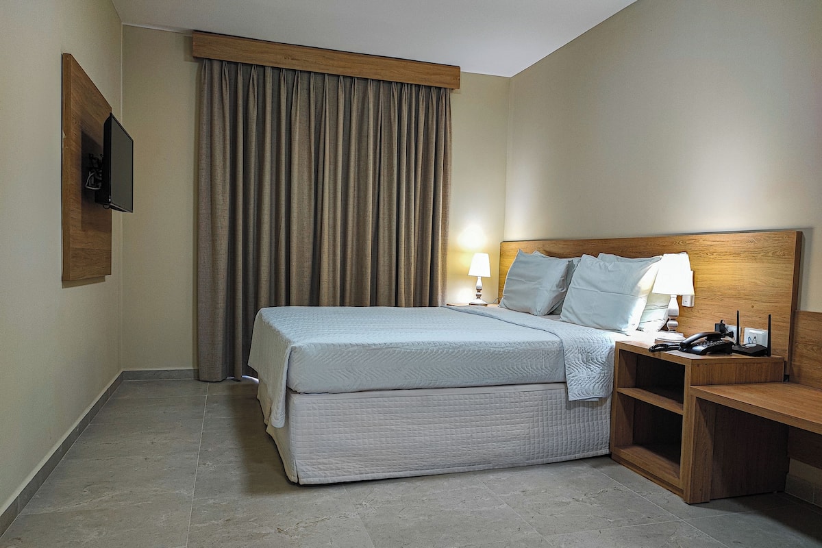 Quarto de hotel com cama, escrivaninha, abajur, telefone e televisão fixada na parede.