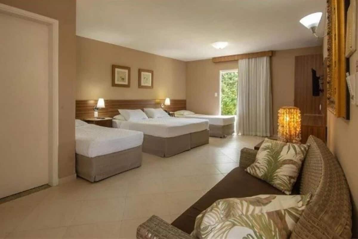 Quarto de hotel com duas camas de solteiro, um sofá, uma luminária e uma janela com cortinas.