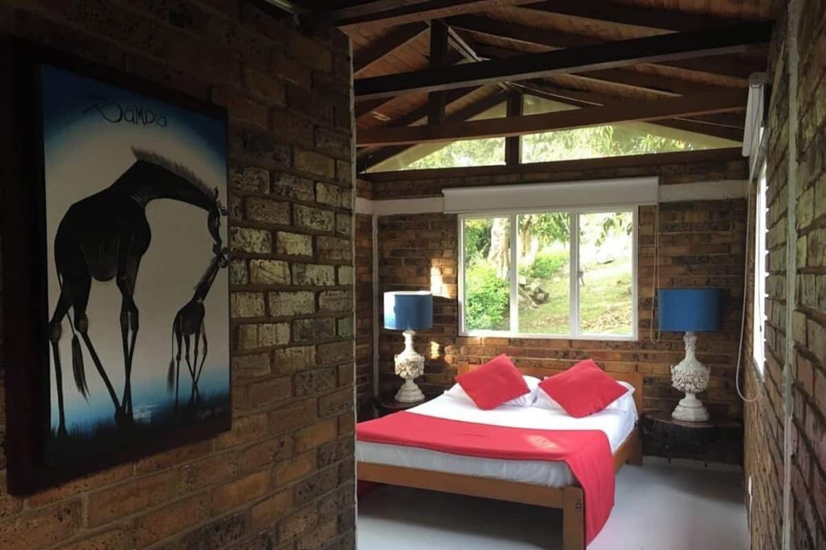 Quarto com parede de tijolos, cama com roupa de cama vermelha, duas luminárias de cabeceira e janela com vista para vegetação.