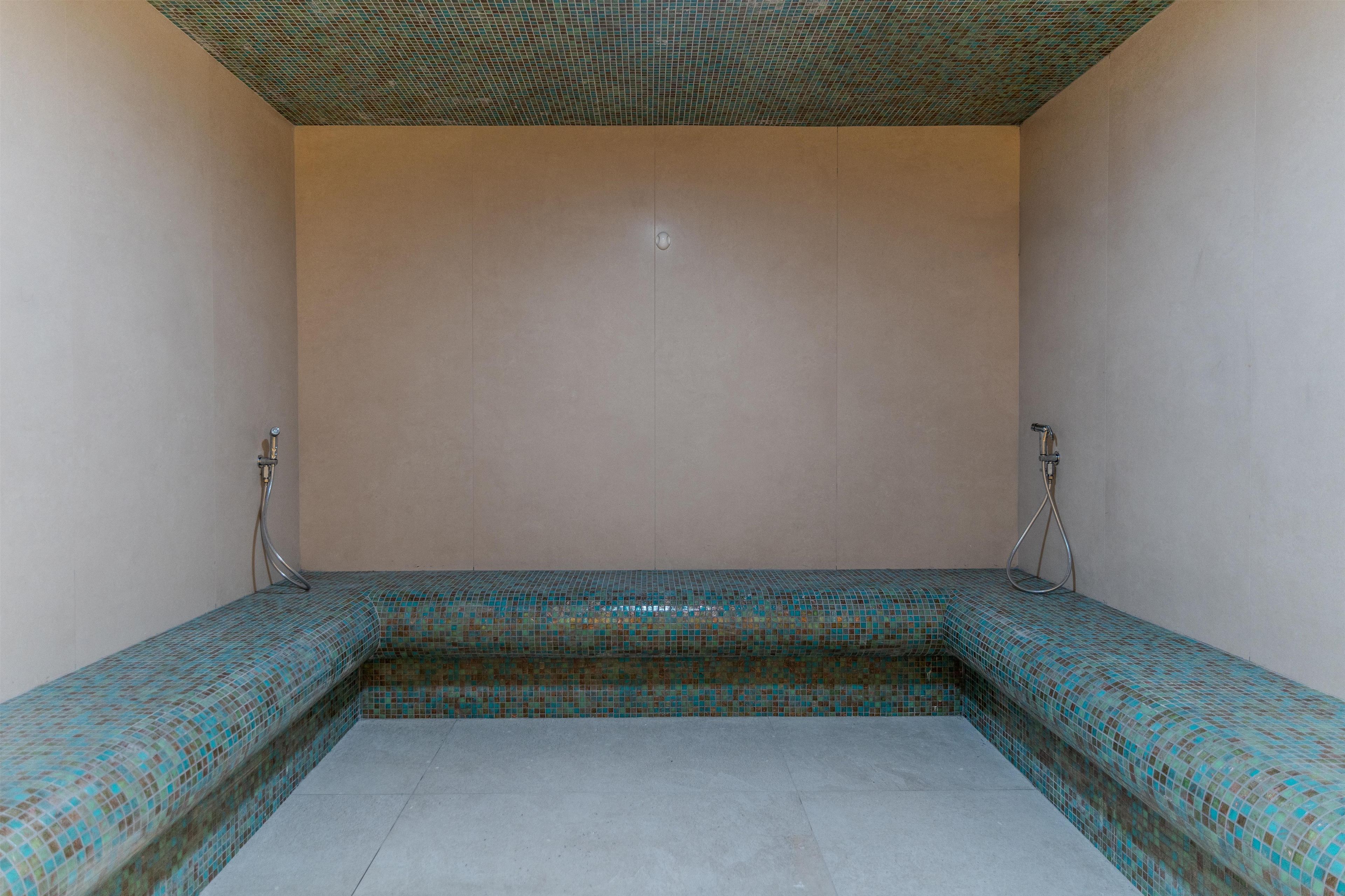 Sala de sauna com piso e paredes revestidos de azulejos, dois chuveiros e um banco.