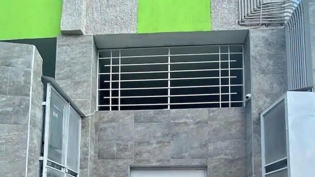 Entrada de um edifício moderno com porta branca, uma pequena janela com grades de metal e piso de concreto.
