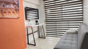 Uma sala de estar com um sofá cinzento, uma mesa pequena branca, uma televisão na parede e uma janela com persianas.