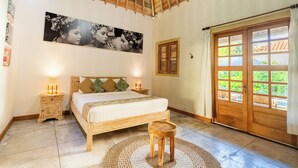 Um quarto com cama, cabeceira de madeira, luminárias de cabeceira, uma moldura grande com uma imagem, uma porta de madeira com painéis de vidro e um tapete de palha no chão.