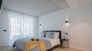Quarto moderno com uma cama grande, uma mesa de cabeceira com um vaso e uma janela com cortinas leves.