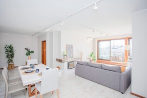 Uma sala de estar moderna com um sofá cinzento, uma mesa de jantar com cadeiras brancas e uma cozinha ao fundo.