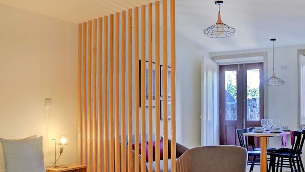 Quarto com cama, divisória de madeira, área de refeições com cadeiras e mesa, e luminária suspensa.