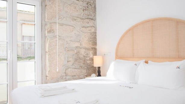 Quarto de hotel com parede de pedra, cabeceira de madeira, cama com lençóis brancos e mesa de cabeceira com abajur.