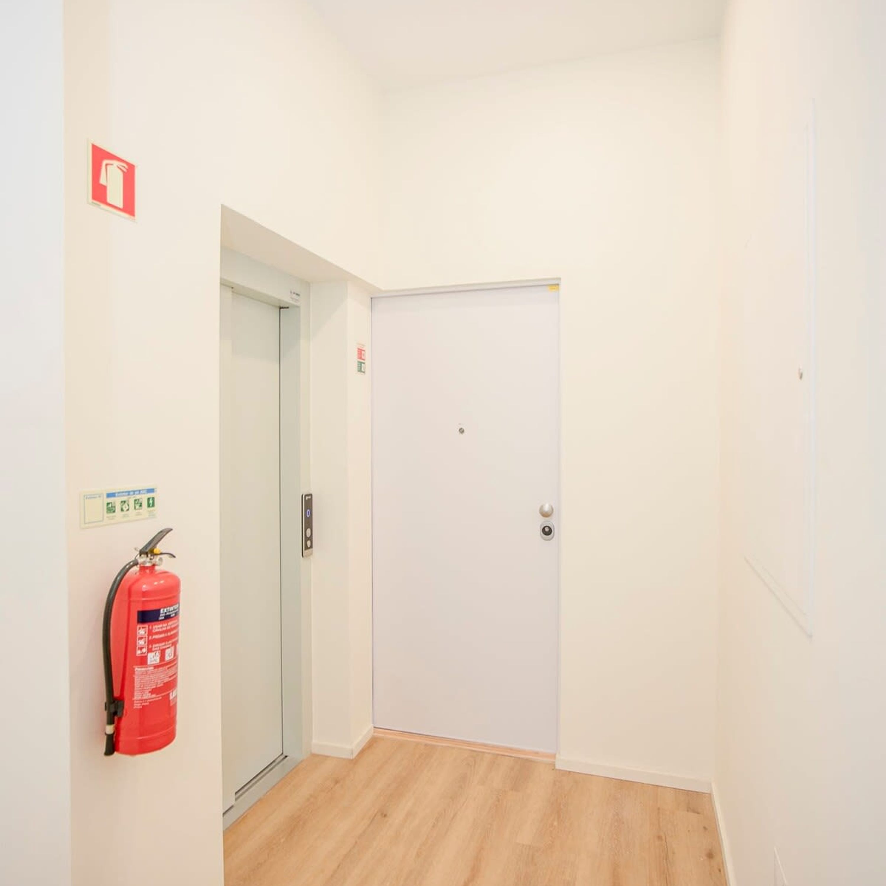 Corredor com extintor fixado na parede, uma porta e uma pequena placa indicando saída de emergência.