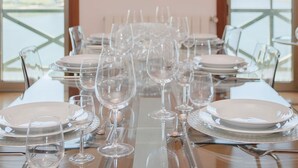 Mesa de jantar posta com vários pratos e copos de vinho, situada em um ambiente com grandes janelas com vista para um corpo d'água.