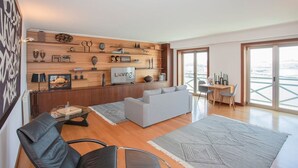 Uma sala de estar moderna com um sofá cinzento, uma poltrona preta, uma parede de madeira com diversos objetos decorativos, uma televisão e uma área de jantar com mesa e cadeiras.