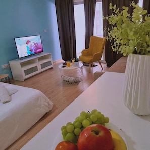 Quarto de hotel moderno com televisão, uma poltrona amarela, um sofá branco e uma mesa com frutas.