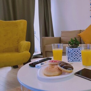 Uma sala de estar com uma poltrona amarela, um sofá, uma mesa de centro com um prato de donuts, dois copos de suco de laranja, um controle remoto e um smartphone.