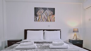 Quarto de hotel com cama grande, duas almofadas brancas, cabeceira preta, pintura de zebras na parede, abajur e pequena mesa com luminária.