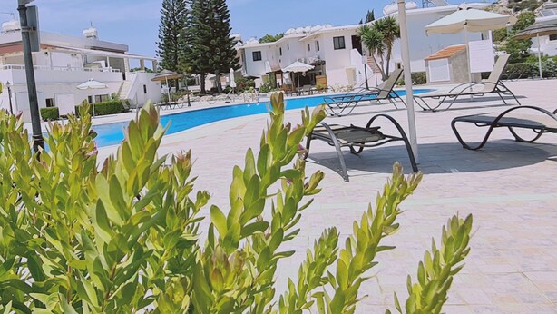 Uma área de piscina com espreguiçadeiras e guarda-sóis, cercada por edifícios brancos e vegetação.