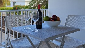 Mesa de metal branca com duas taças de vinho e uma garrafa, uma cesta de frutas e uma cadeira branca numa varanda.