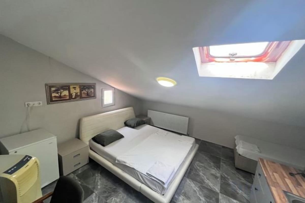 Quarto compacto com cama, mesa de cabeceira e uma pequena escrivaninha com cadeira. Há um claraboia e um radiador na parede.