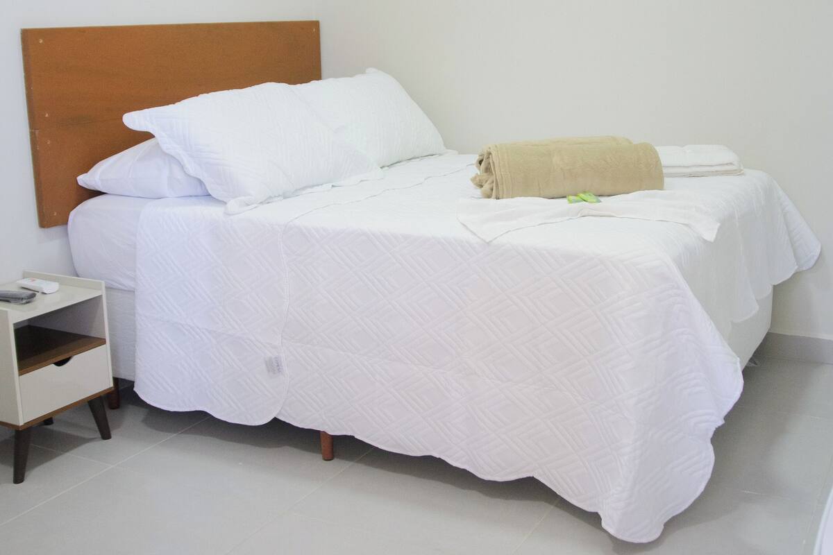 Uma cama bem arrumada com roupa de cama branca e toalha bege, cabeceira de madeira, mesa de cabeceira pequena e parede branca lisa.