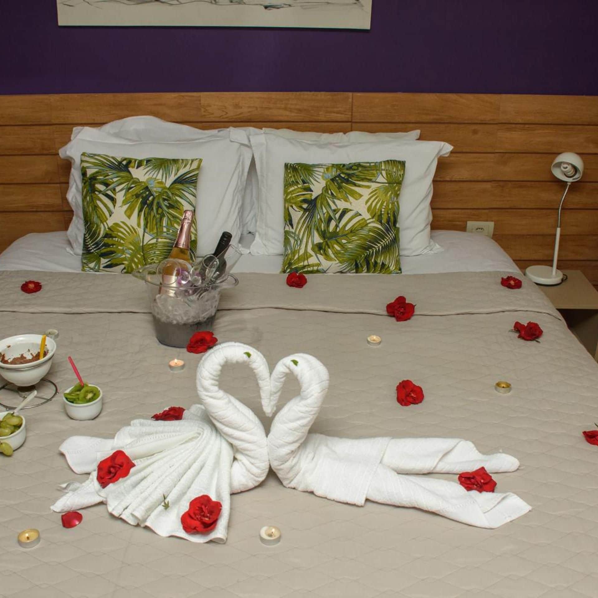 Quarto de hotel com uma cama, duas almofadas com padrão de folha tropical, mesa de cabeceira com luminária, garrafa, copos e uma tigela com comida.