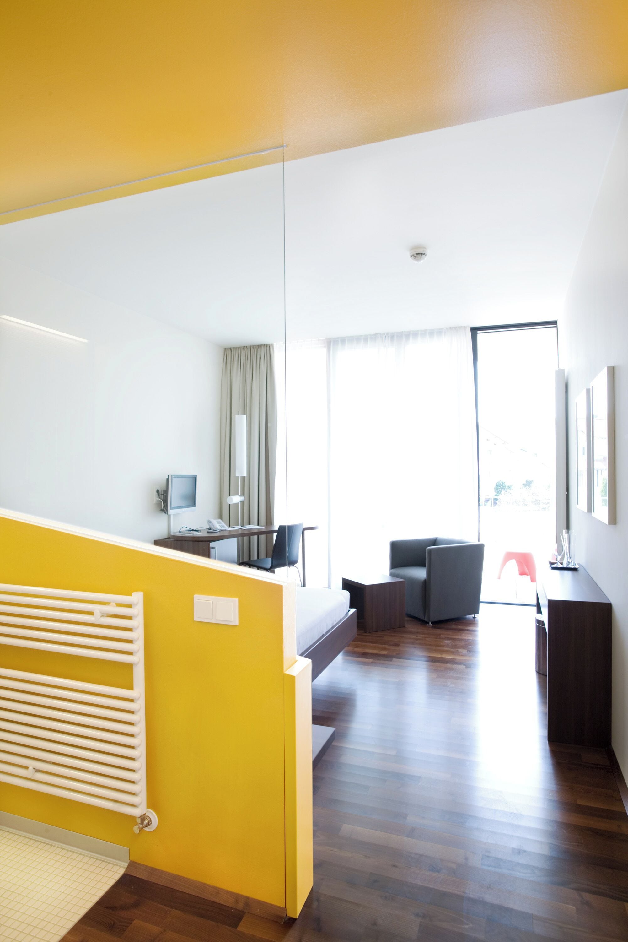 Quarto de hotel moderno com parede amarela de destaque, piso de madeira, escrivaninha com computador, sofá e uma janela grande com cortinas.