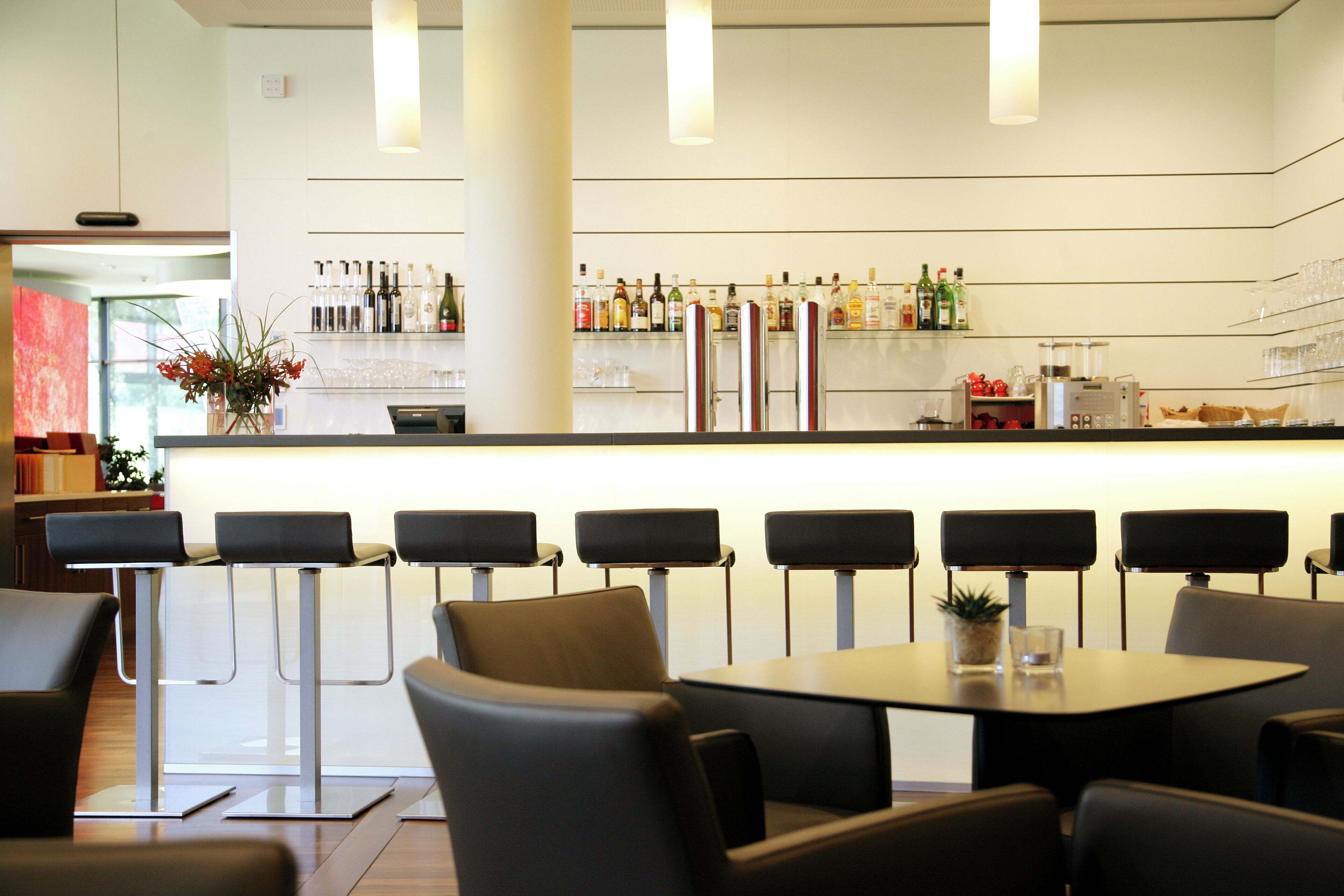 Uma área de bar moderna com um balcão extenso, diversas opções de assentos e uma variedade de garrafas e copos nas prateleiras.