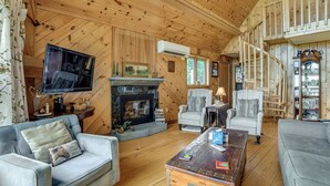 Interior acolhedor de uma cabana de madeira com lareira, dois sofás, uma televisão e uma escada de madeira.