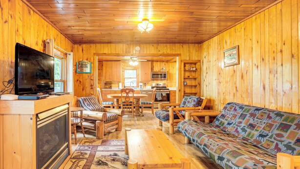 Interior acolhedor de uma cabana, com chão de madeira, sofá, mesa de jantar e televisão.