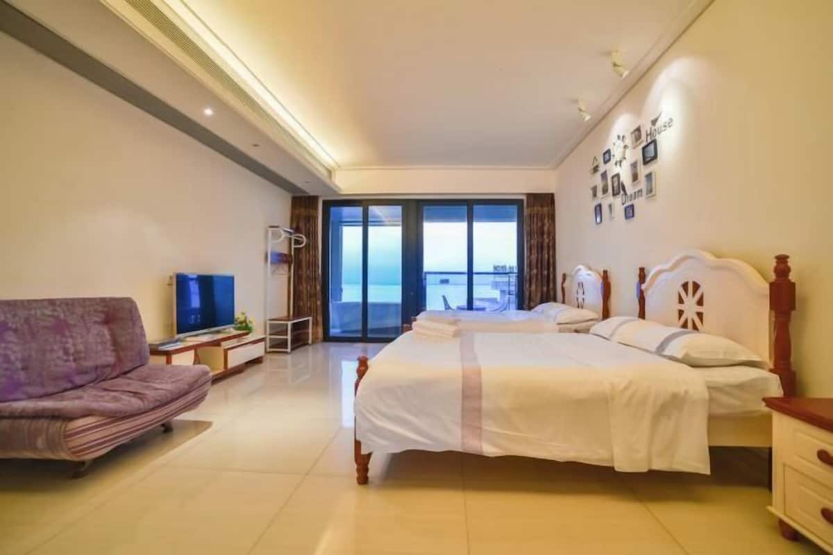 Quarto de hotel com cama, sofá, televisão e vista para o mar através de uma porta de vidro.