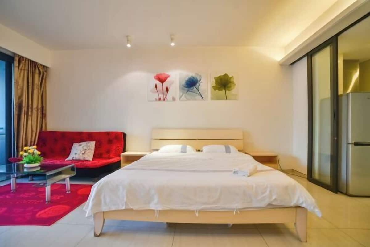 Quarto de hotel moderno com uma cama grande, um sofá vermelho, uma mesa pequena com uma planta e obras de arte na parede.