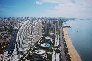 Um edifício alto com um design curvo e distinto, rodeado por construções menores e uma orla marítima.